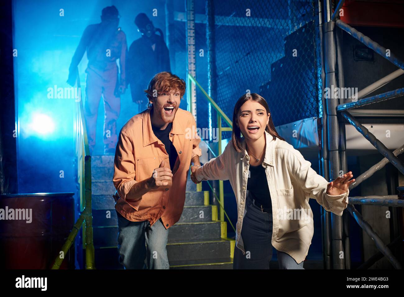 scared man and woman running from freaky people in gas masks and orange ppe suits in escape room ...