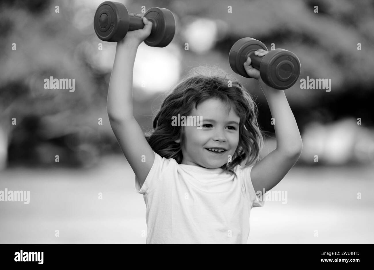 Happy little excited boy doing exercises with dumbbells in green park ...