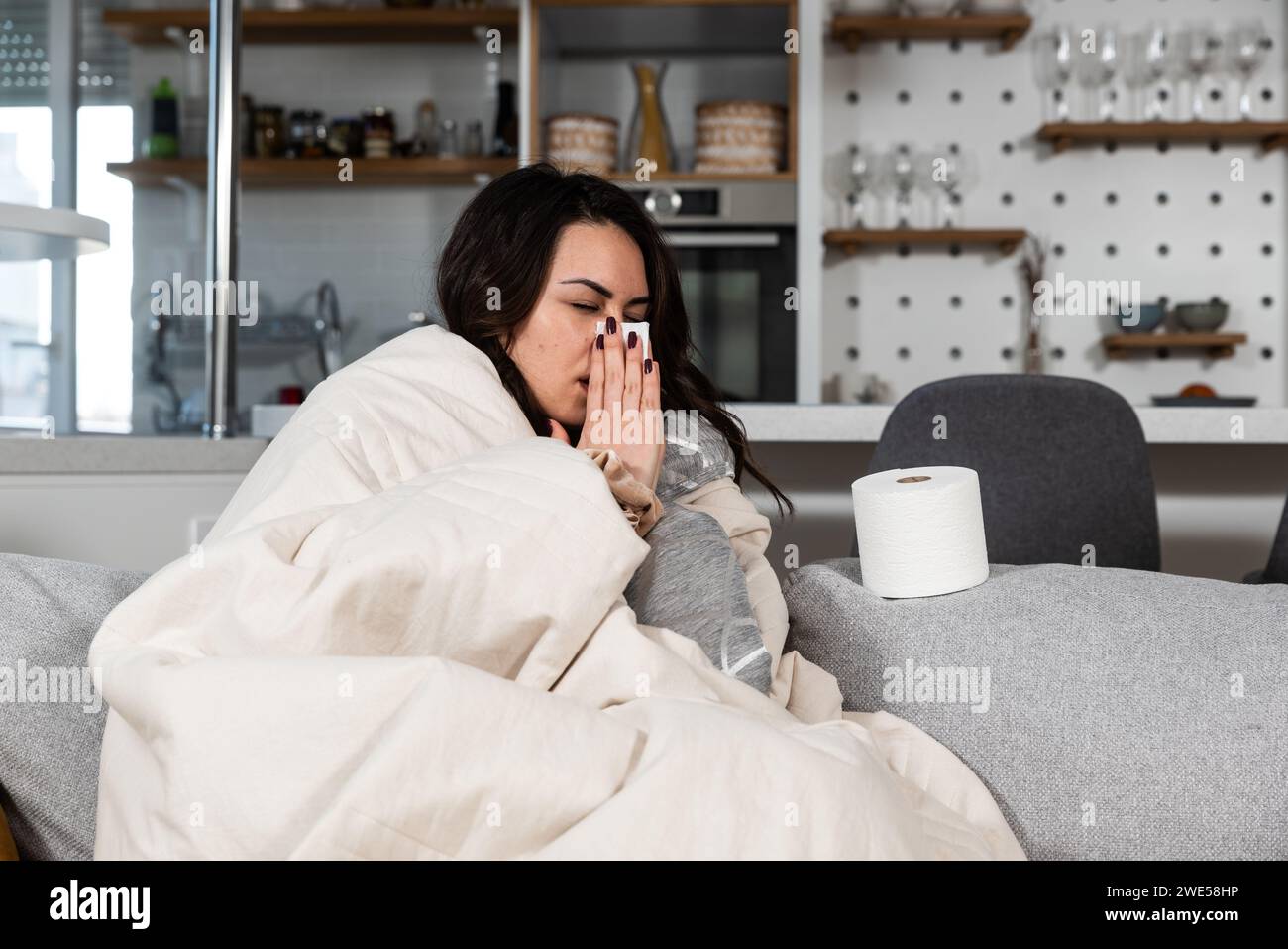 Young sick woman wrapped in a blanket wipes her nose with paper tissues ...