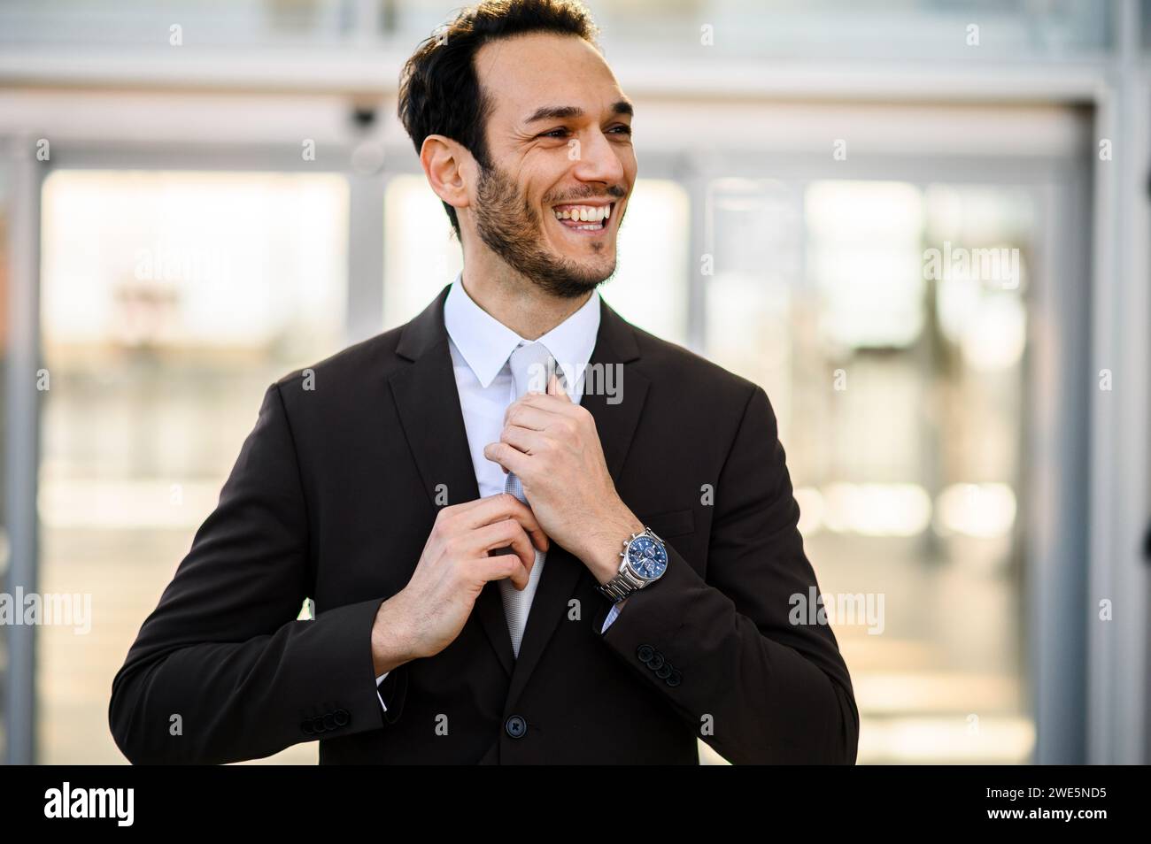 Happy young man getting ready for work, fixing his tie with a modern building backdrop Stock ...