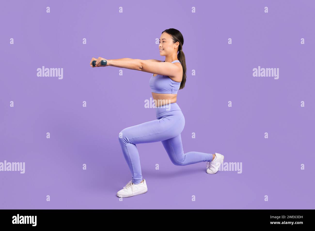 Asian woman in lunge holding dumbbell with extended arms on purple ...