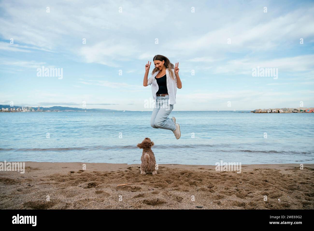 Playful young woman gesturing peace sign and jumping near poodle dog at ...