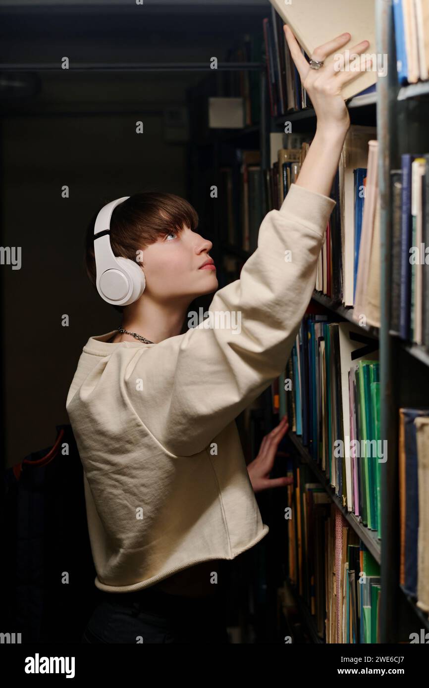 Young student wearing wireless headphones and taking book from top ...