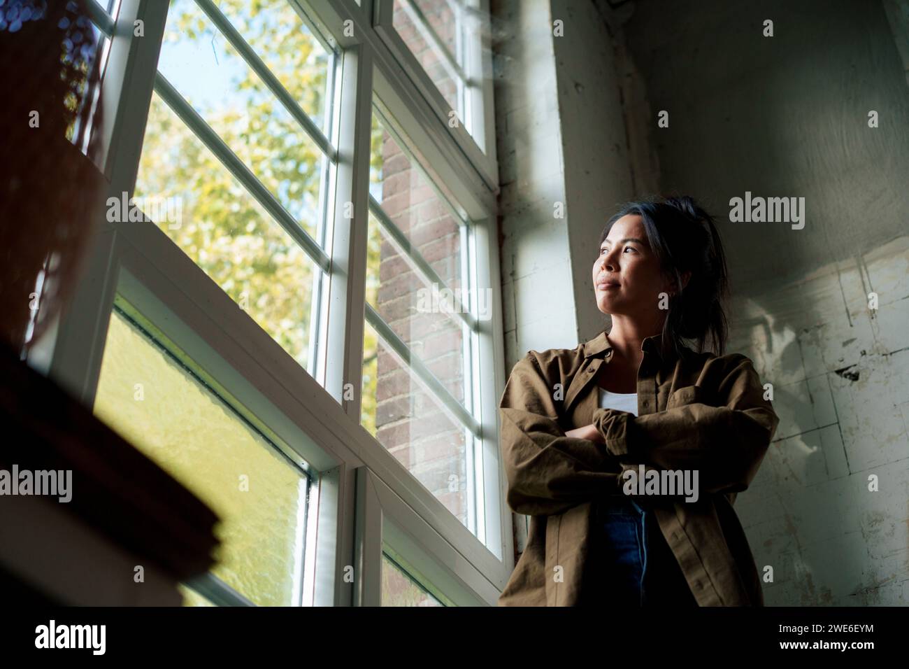 Thoughtful woman with arms crossed looking out through window at home ...