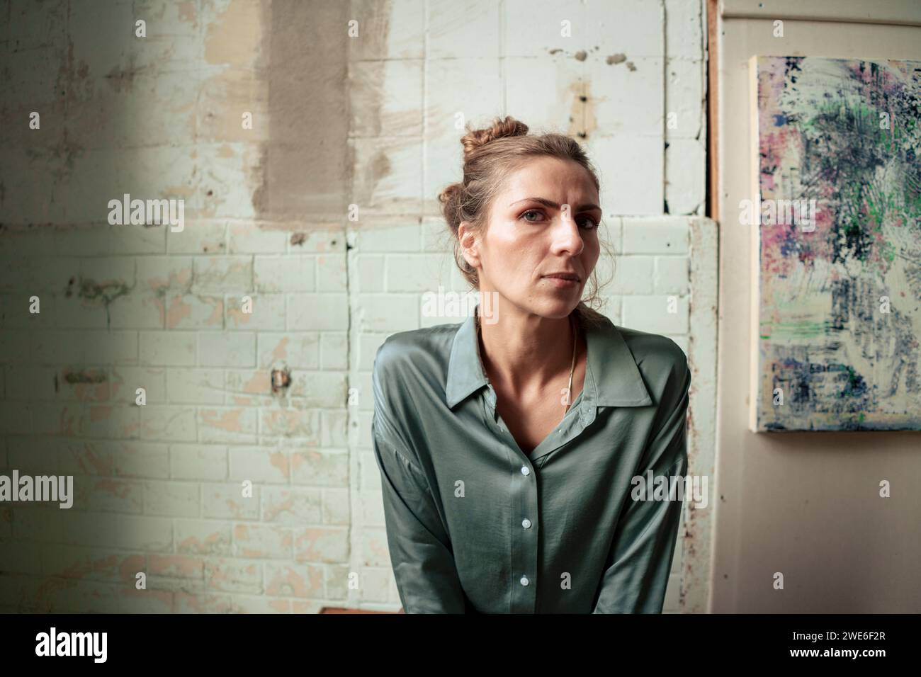 Confident artist sitting by painting in front of brick wall at studio ...