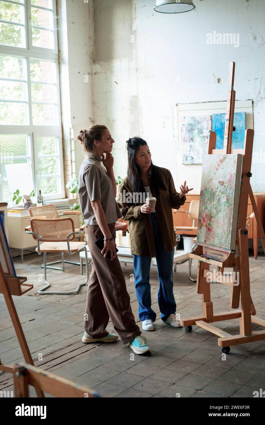 Confident women discussing over painting on easel in art studio Stock ...