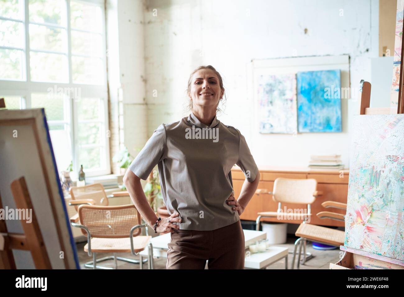 Smiling painter standing with arms akimbo by painting on easel in ...