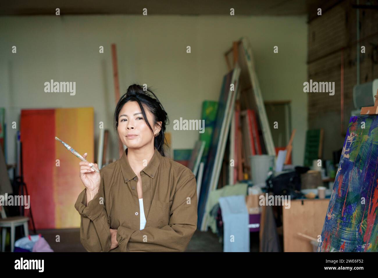 Confident painter holding paintbrush standing by painting at workshop ...