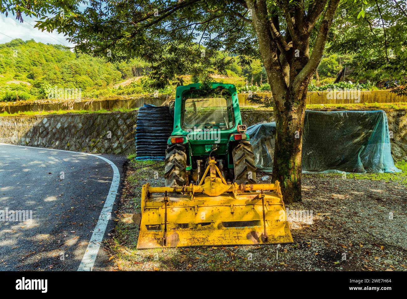 Green tractor with yellow cutting attachment parked under a large shade ...