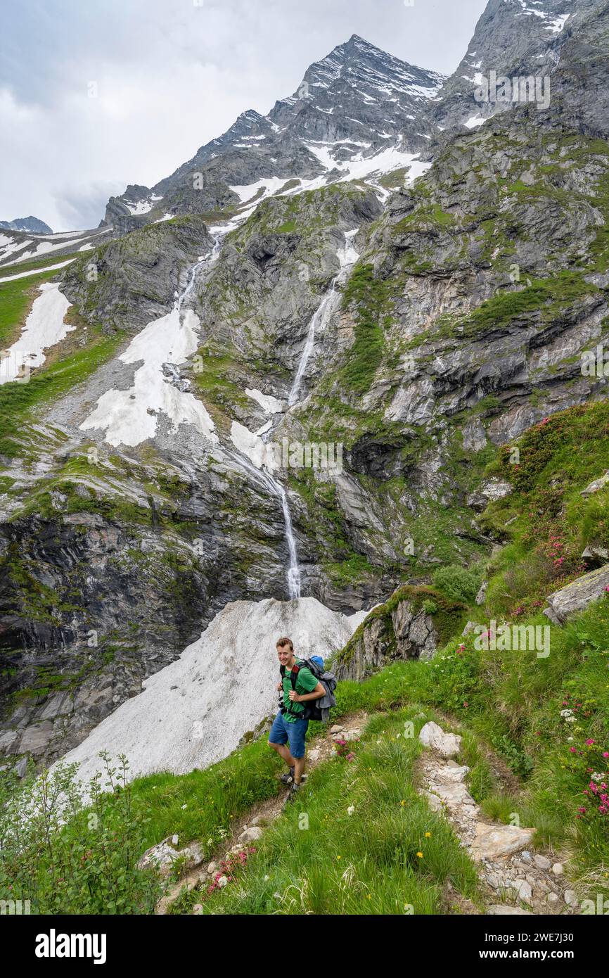 Mountaineer on a hiking trail, mountain landscape with small waterfall and rocky mountain peak ...