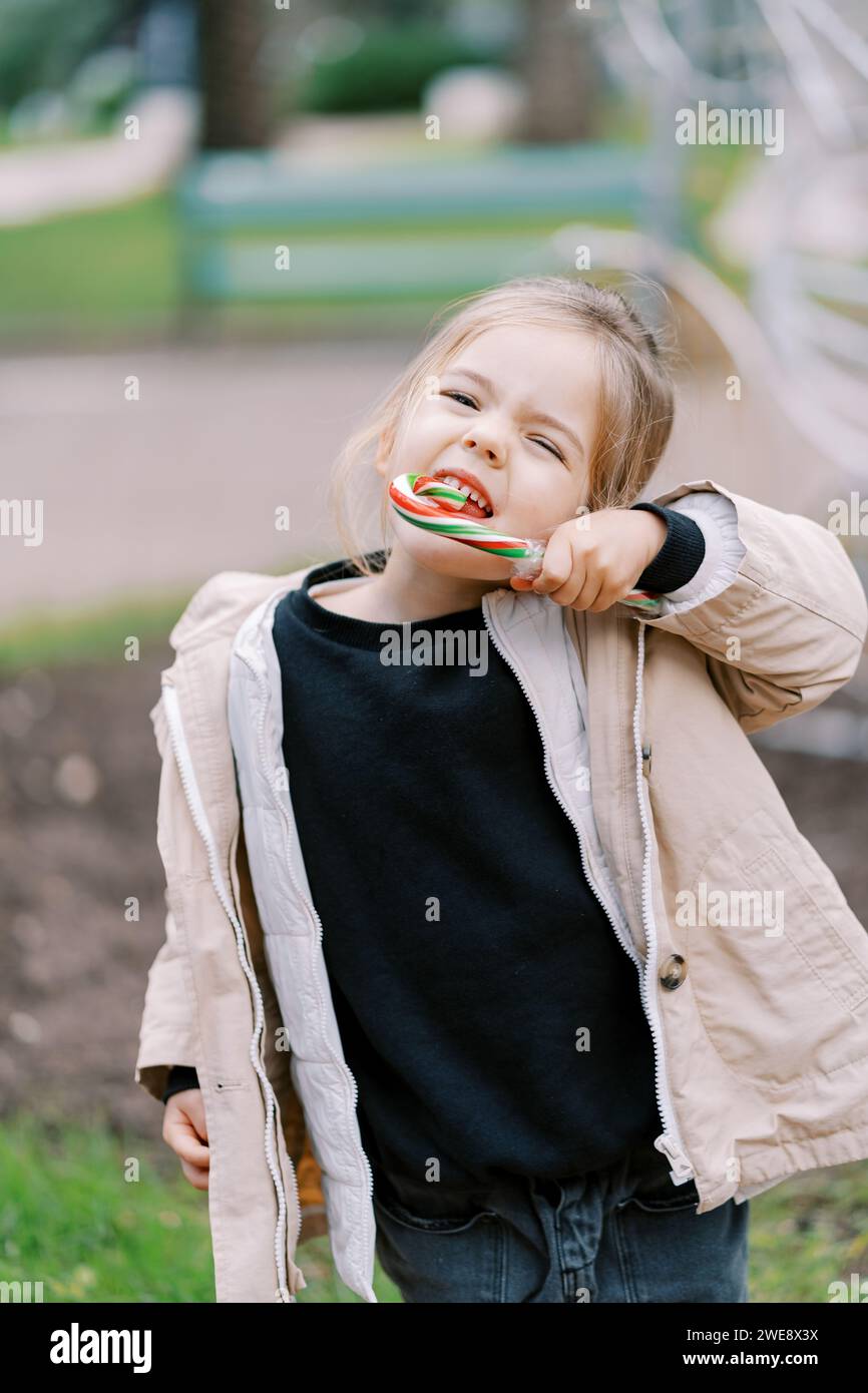 Little girl chews on a striped candy cane while standing in the park ...