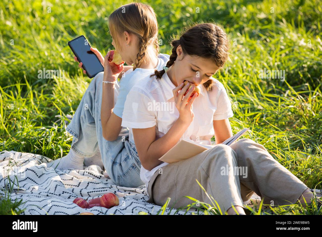 Two Teenage Girls Spend Time On Green Grass Lawn In Park Together One Of Them Eating An Apple