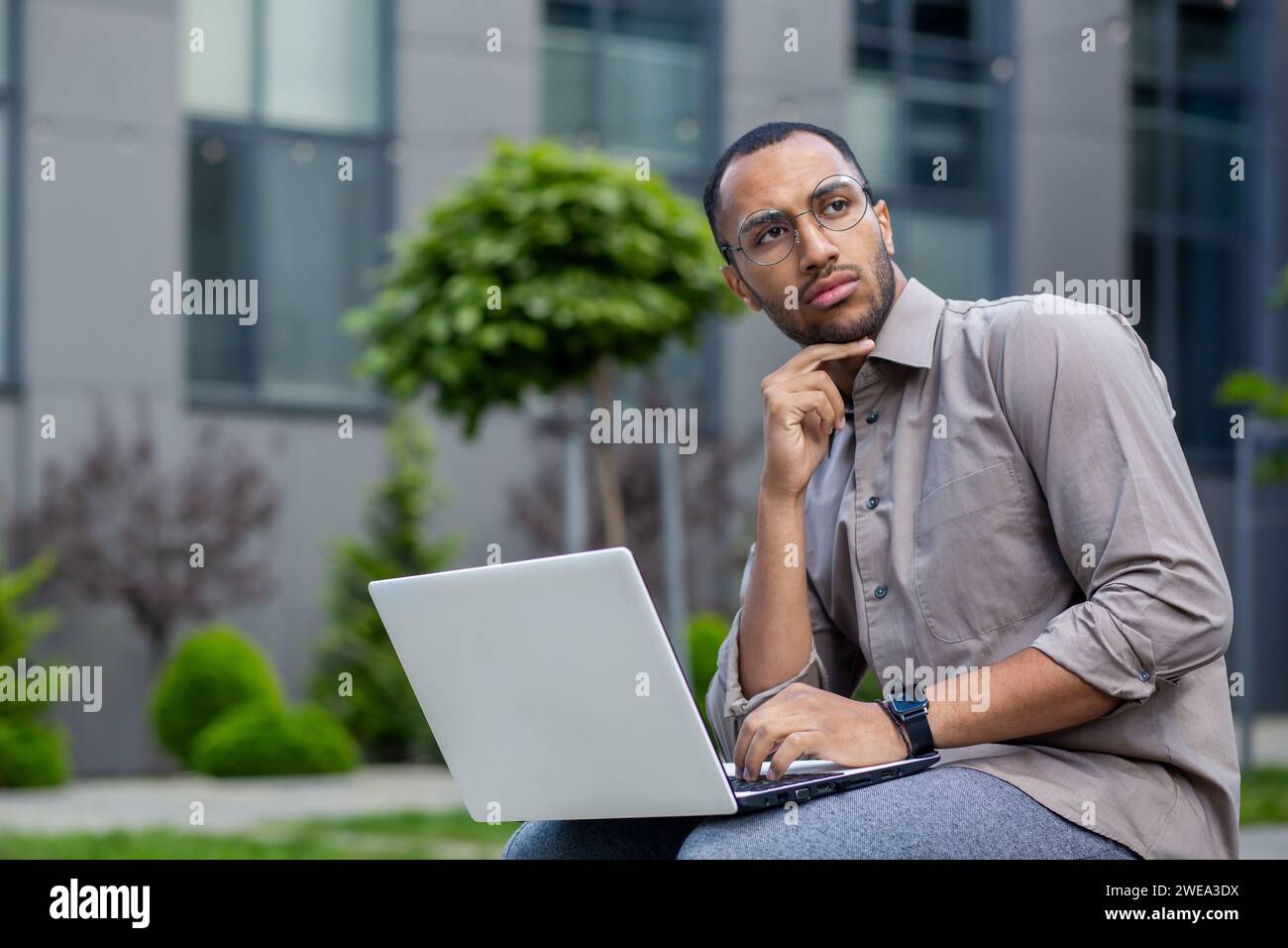 Young businessman thinking sitting outside office building with laptop ...