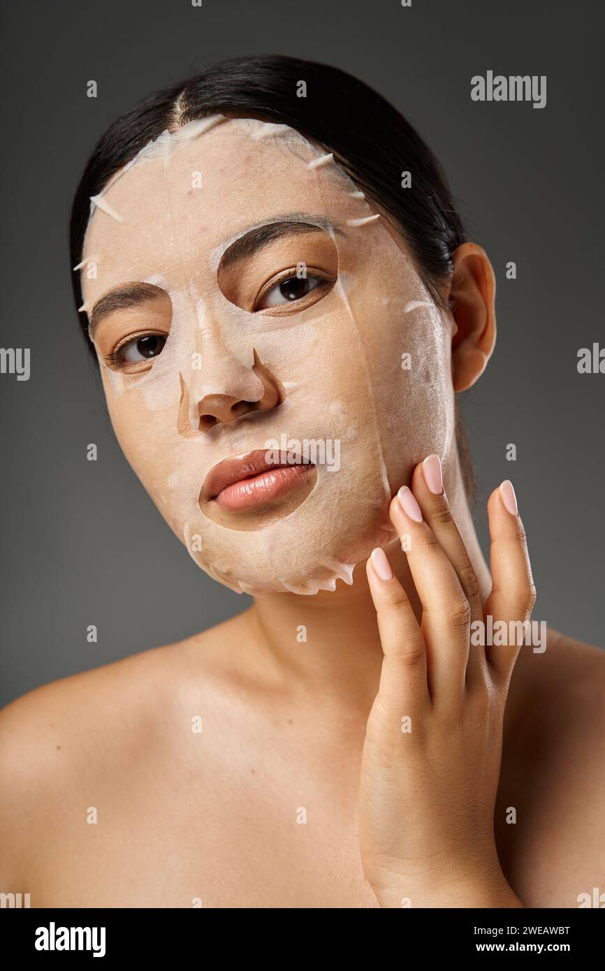 brunette young asian woman touching face with moisturizing sheet mask ...