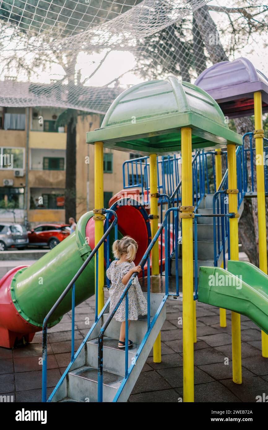 Little girl climbs the steps of a colorful slide, holding the handrail ...