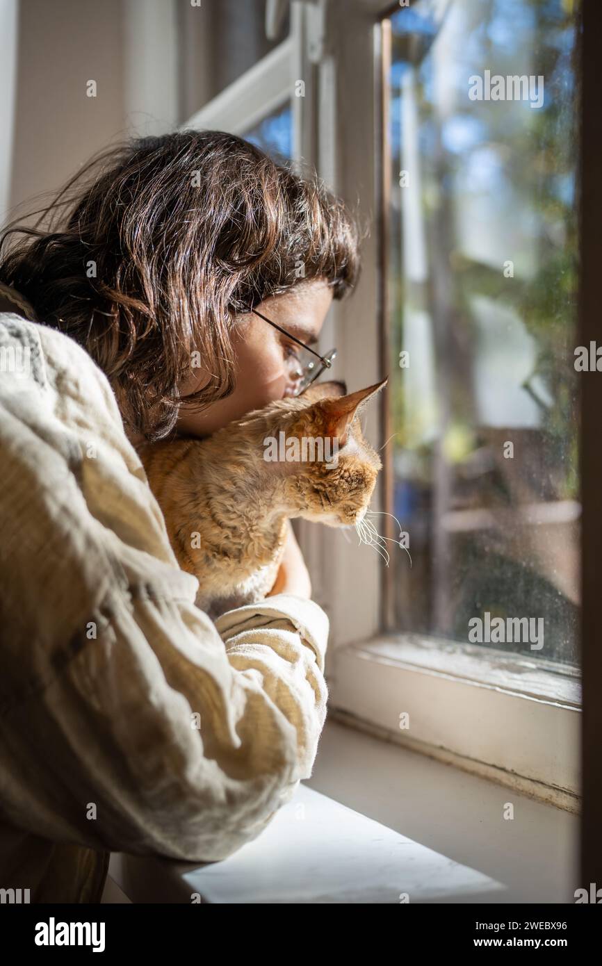 Caring pet lover cuddling beloved breed cat Devon Rex sitting on windowsill looking at window ...