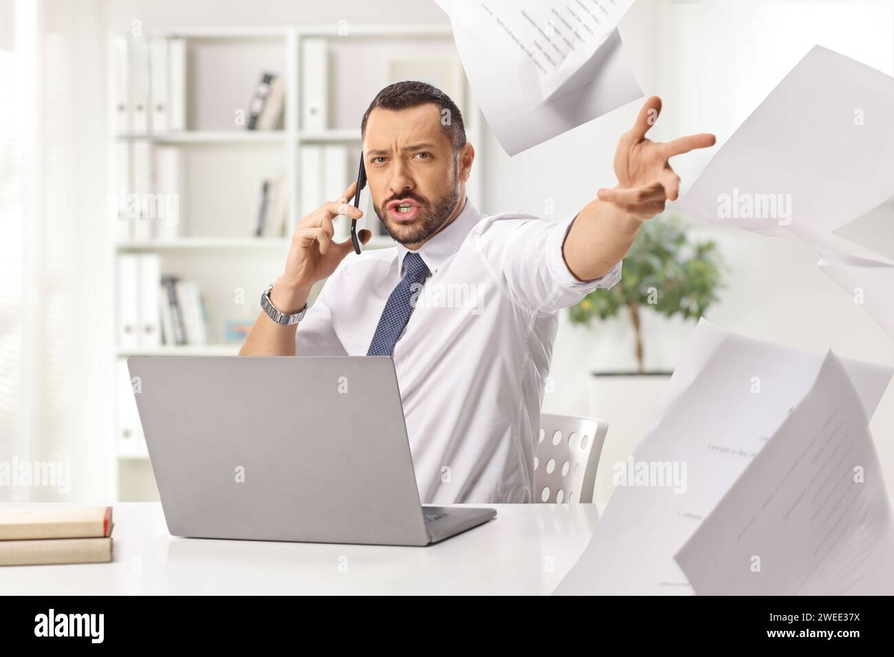 Angry boss at a desk throwing papers and making a phone call in the ...