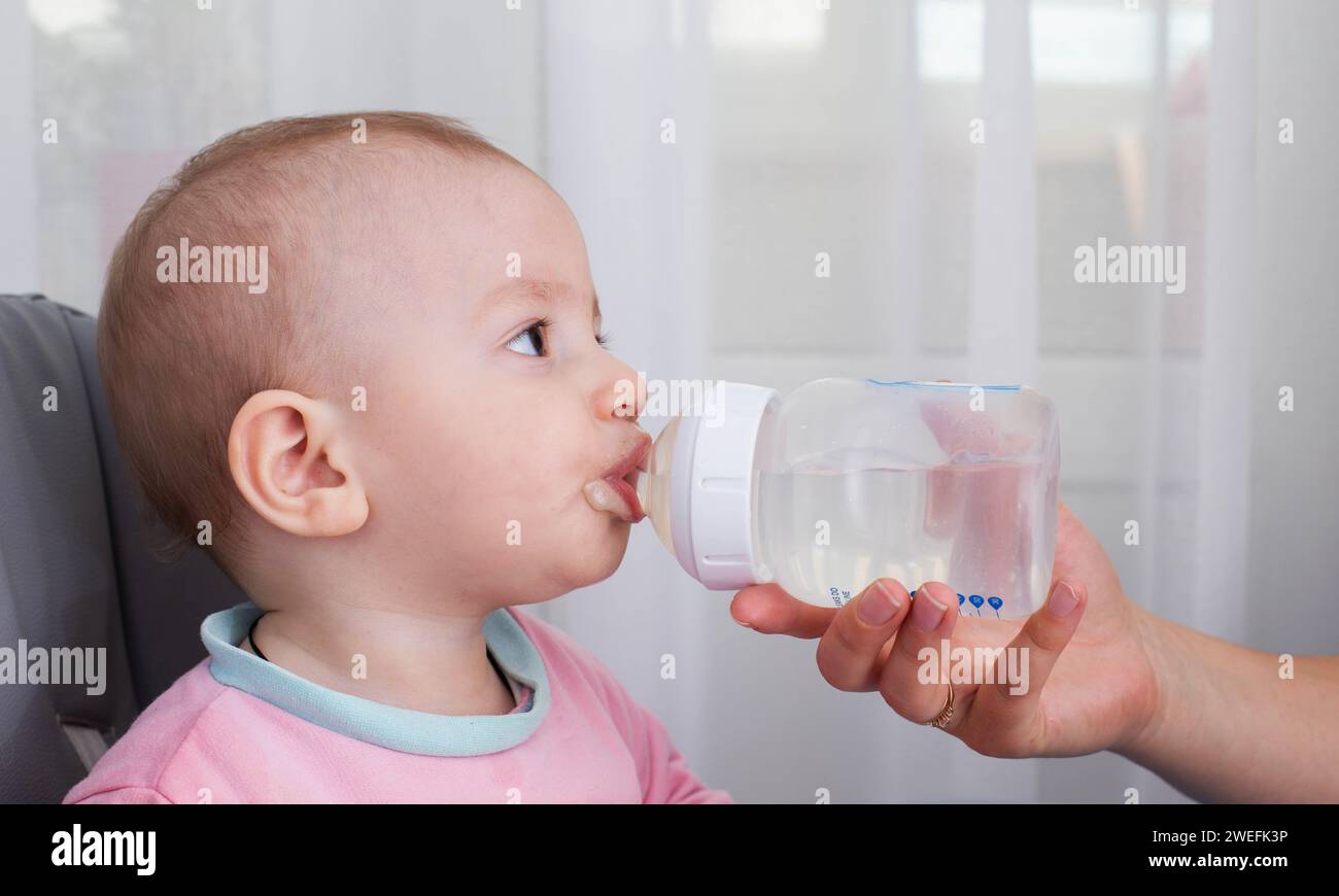A mother's hand gives water to a baby to drink from a bottle while ...