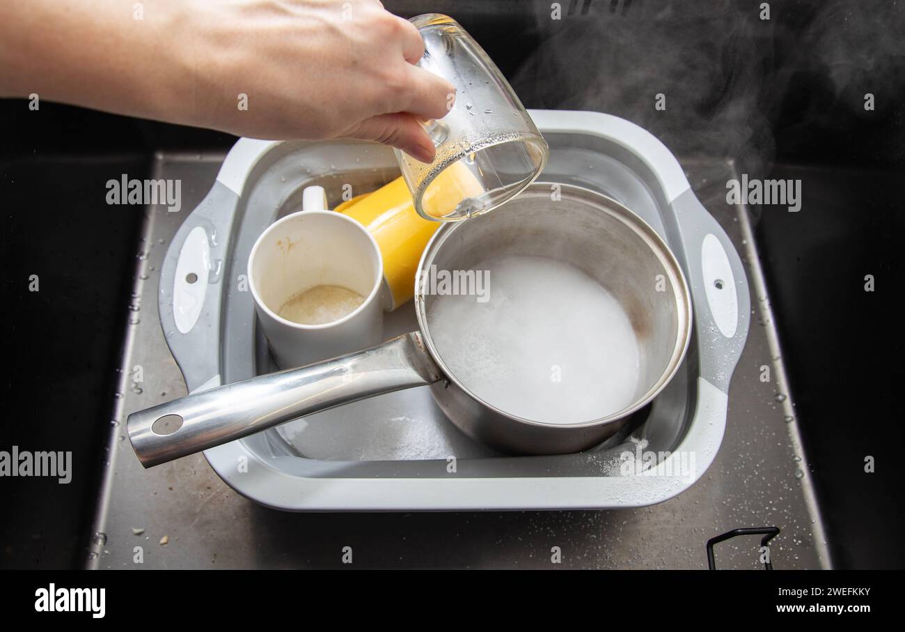 A woman's hand pours hot water into a basin with dirty dishes and ...