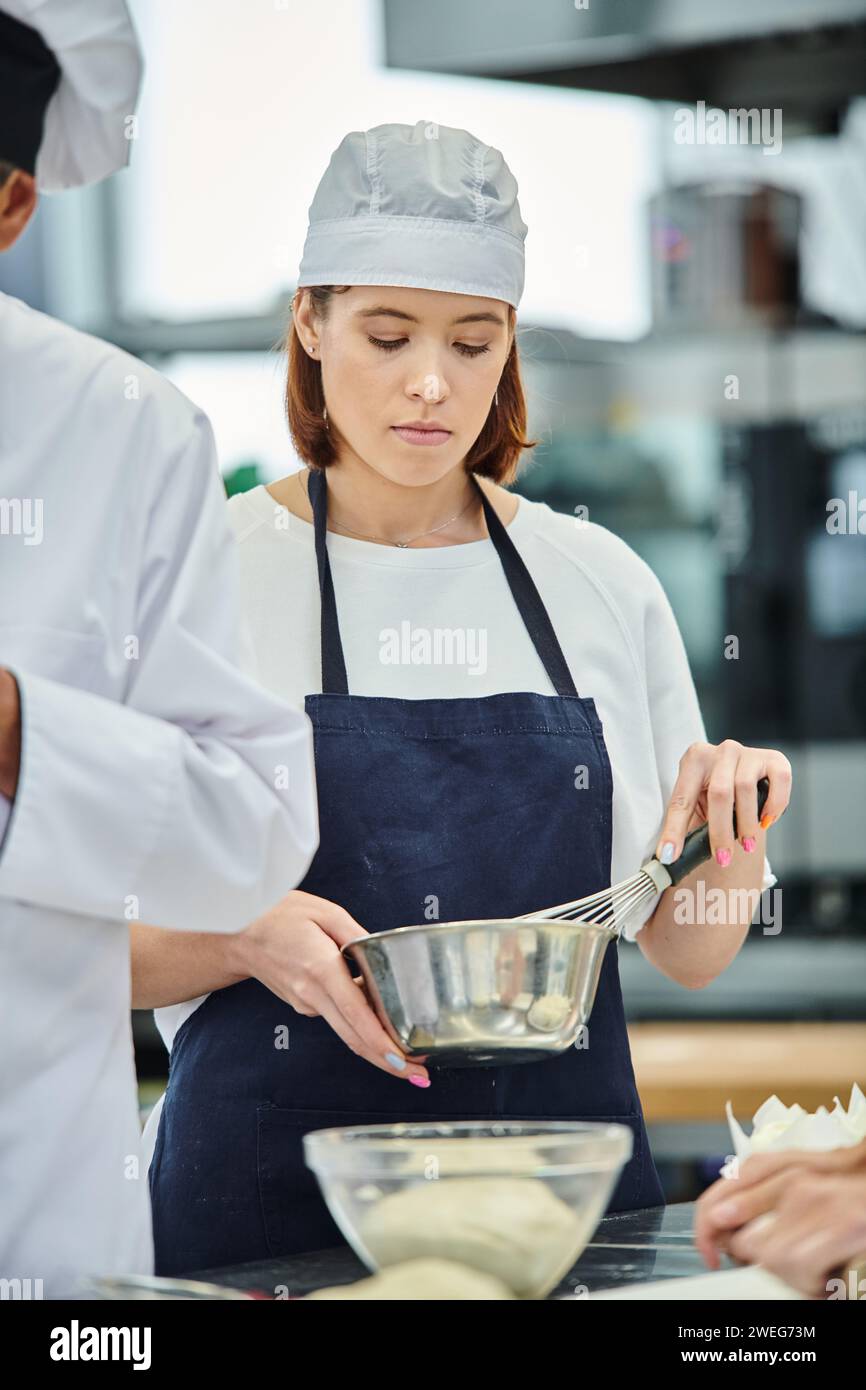 attractive young female chef in toque and apron whisking dough before ...