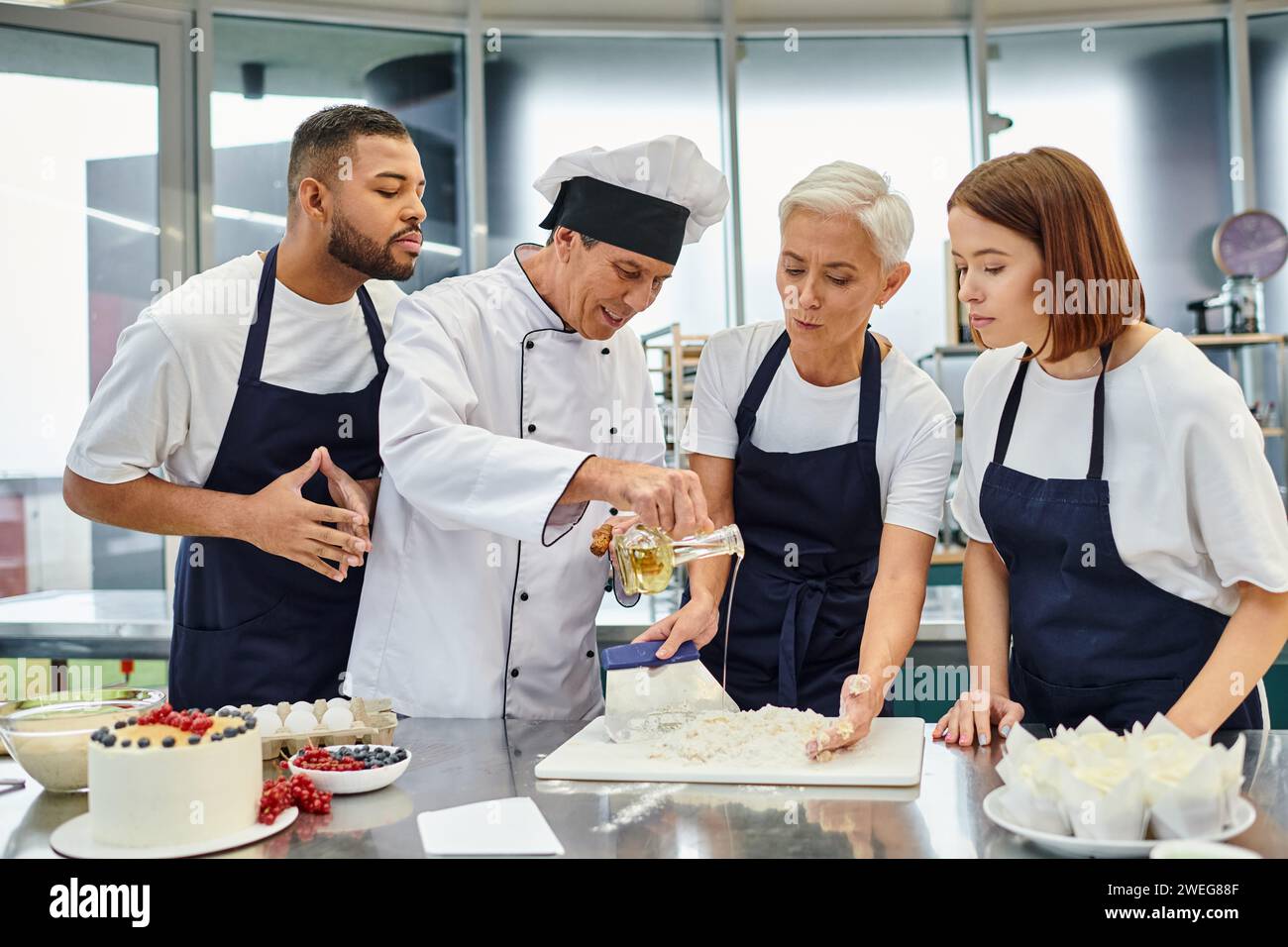 joyful multiracial chefs in blue aprons watching chief cook pouring oil ...