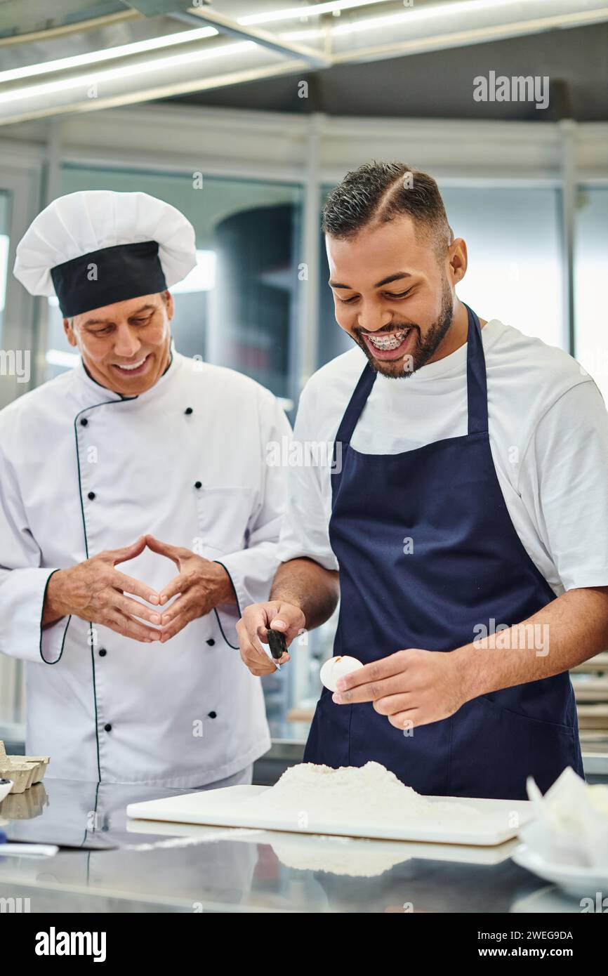 cheerful young african american chef in blue apron breaking egg next to ...
