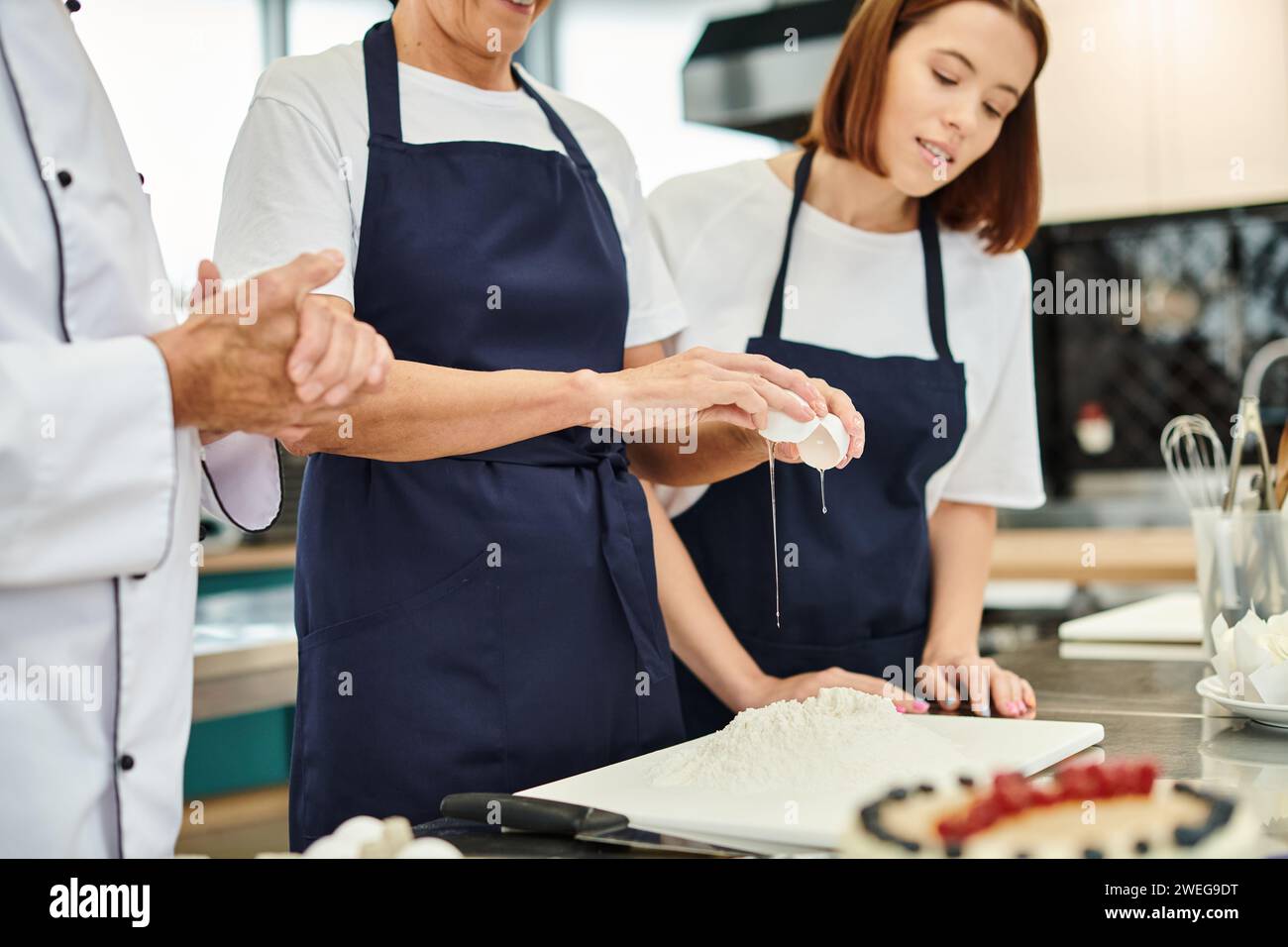 cropped view of mature female chef breaking egg next to her young ...