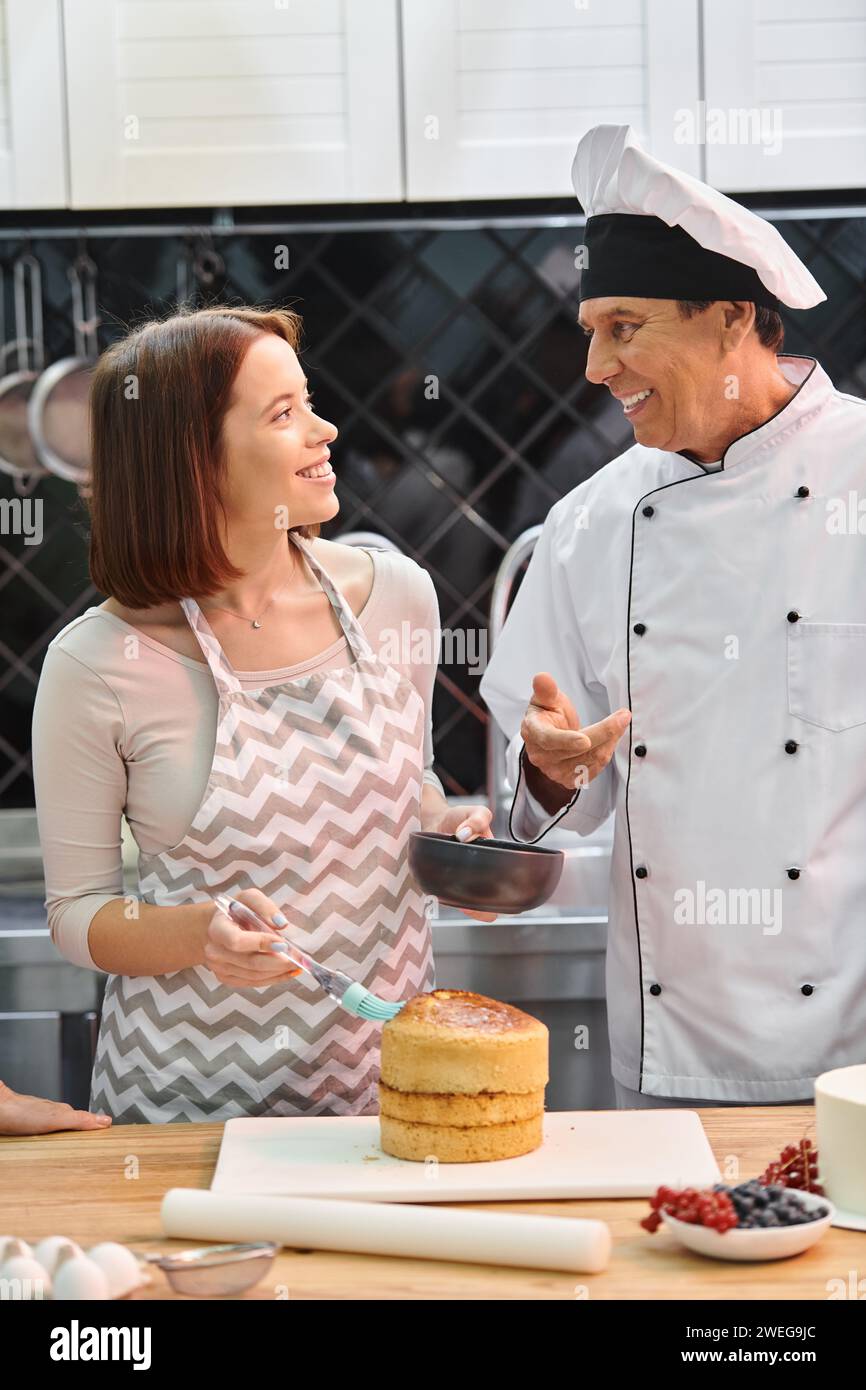 jolly mature chef in white hat looking at young joyful student while ...