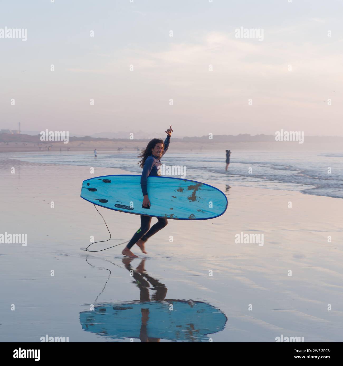 Male surfer in wet suit with long hair smiles with his arm in the air ...