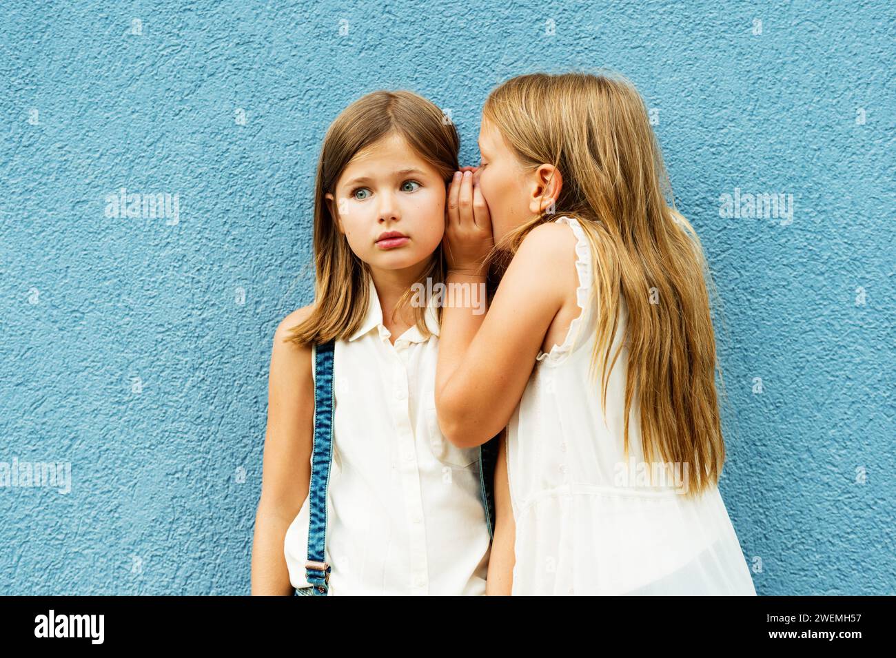 Two cute little girls sharing secrets, standing in front of blue wall ...