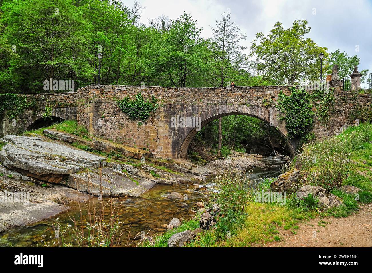 A charming old stone bridge arches over a clear stream surrounded by ...