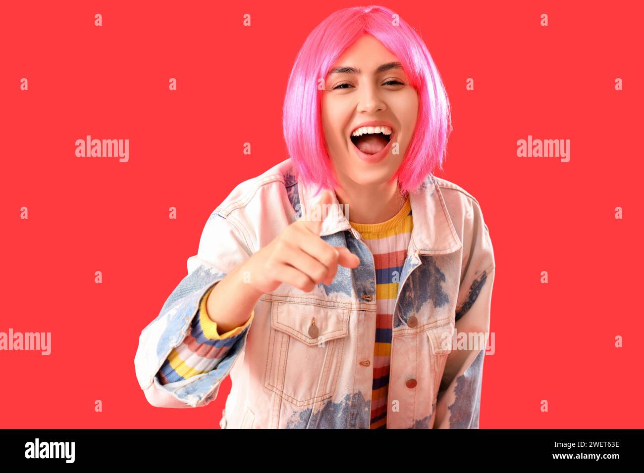 Laughing young woman in wig pointing at viewer on red background ...