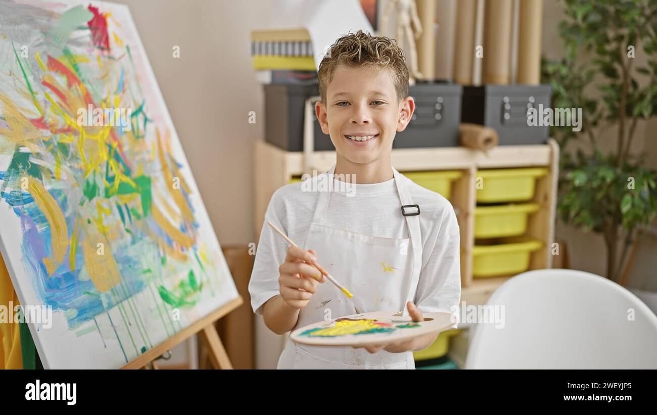 Adorable blond boy artist, confidently holding paintbrush and palette ...