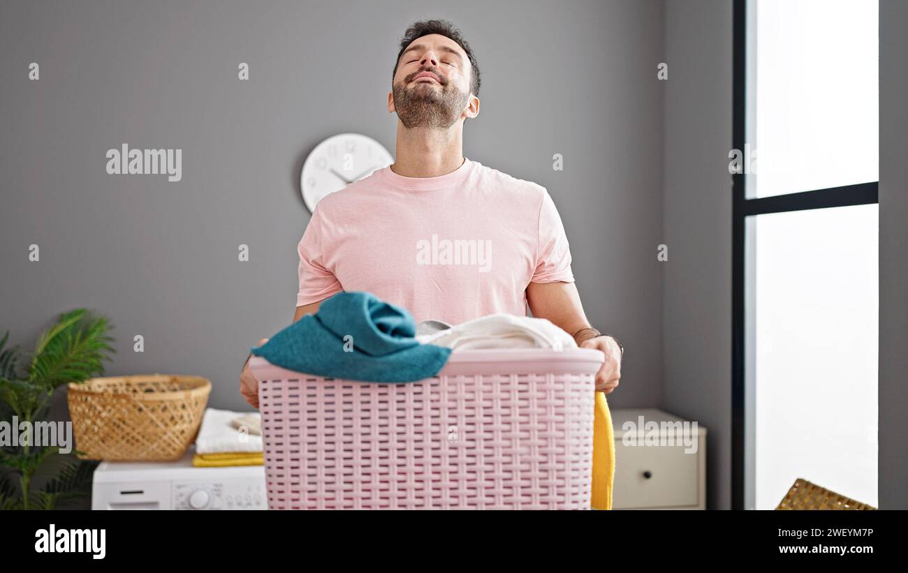 Young hispanic man smiling confident smelling clean clothes on basket ...