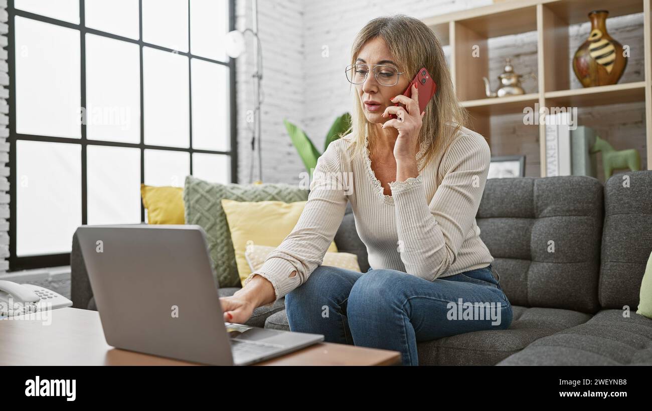 A focused middle-aged woman multitasks using a phone and laptop in a ...