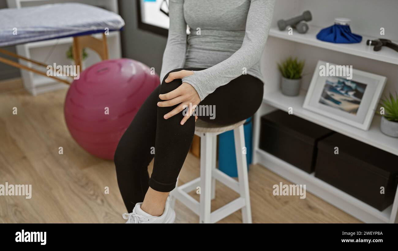 Hispanic young woman in physical therapy clinic holding knee, sitting ...