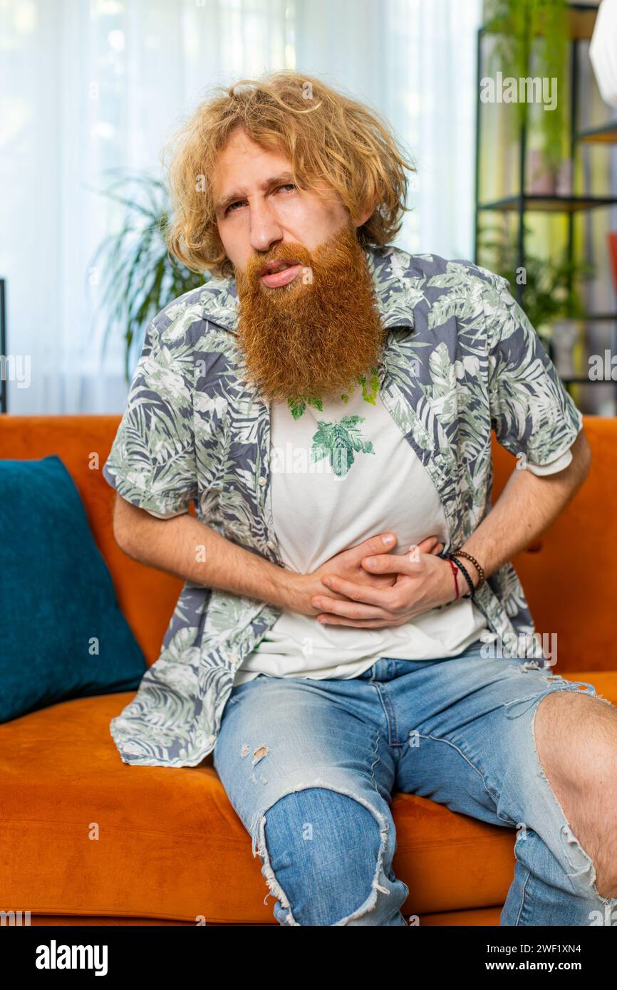Caucasian man sits on sofa feeling sudden strong abdominal stomach ache ...
