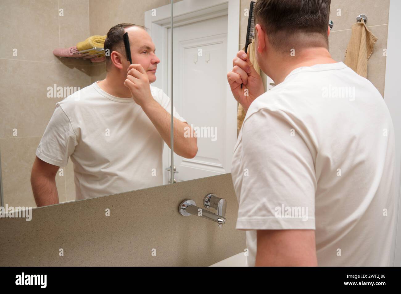 A man combs his hair with a comb in the bathroom in front of the mirror ...