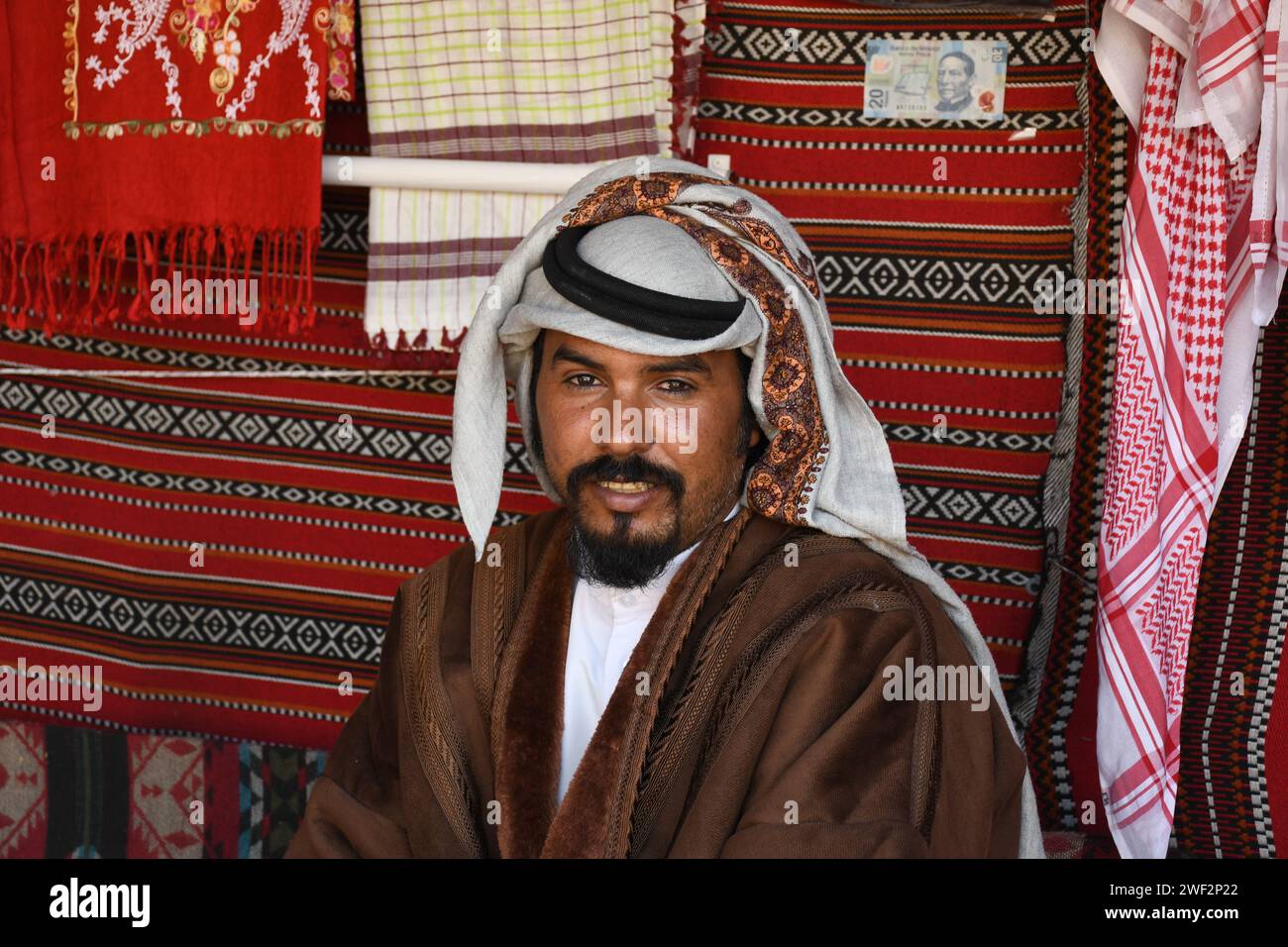 Man in traditional attire sits beside shop displaying various textiles ...