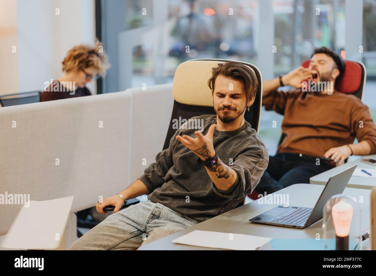 Two tired male colleagues in a modern office setup stretch and yawn ...