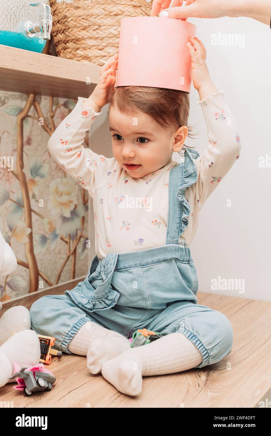 Little baby girl sitting on the table putting paper bucket on her head ...