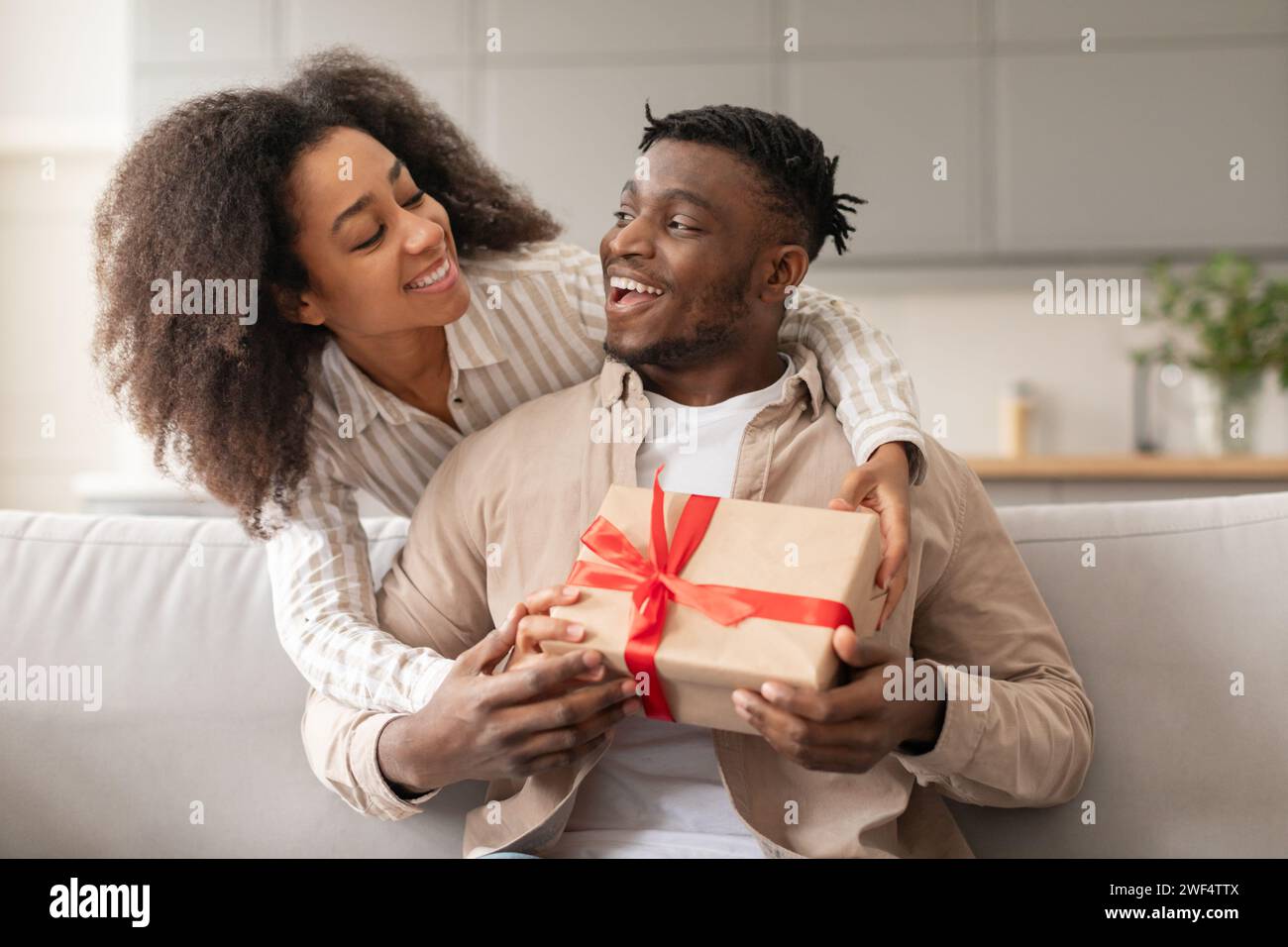 Cheerful black wife surprising her husband giving present box indoors Stock Photo - Alamy