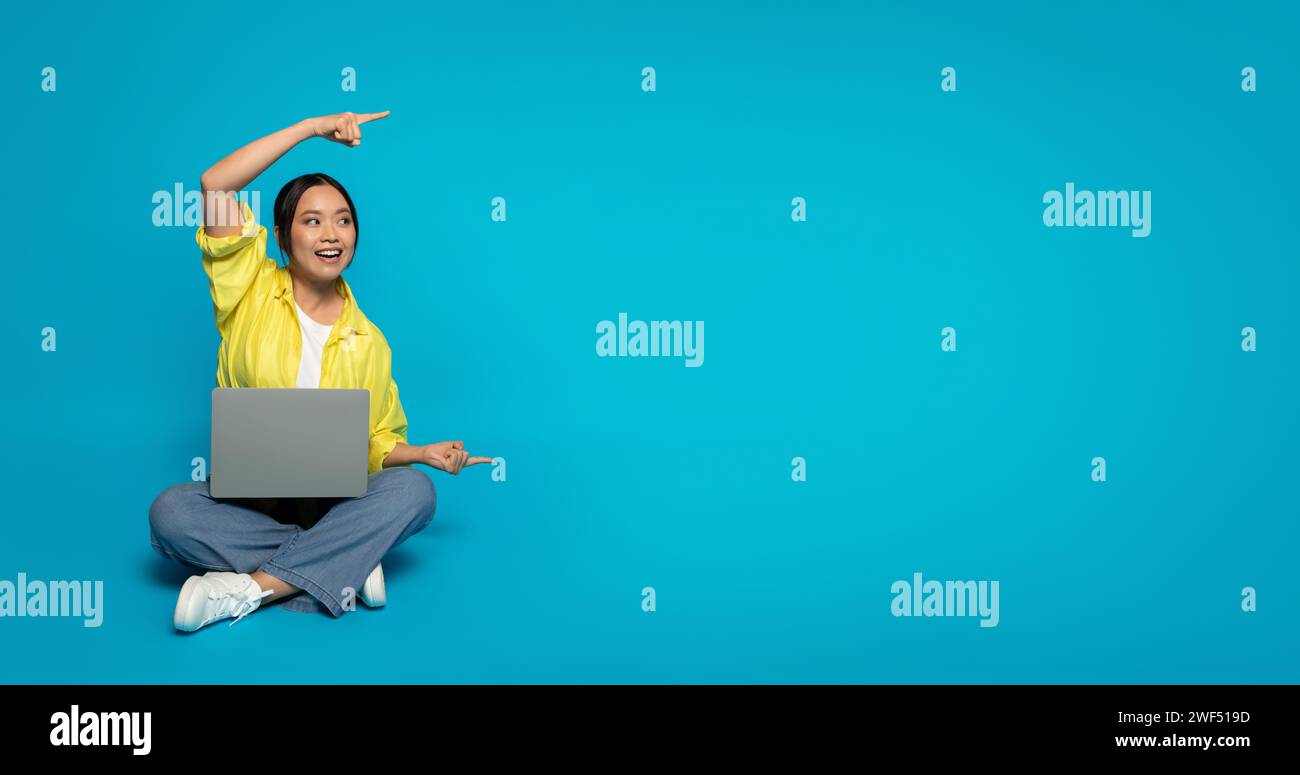 Creative woman sitting cross-legged with a laptop, pointing upwards to ...