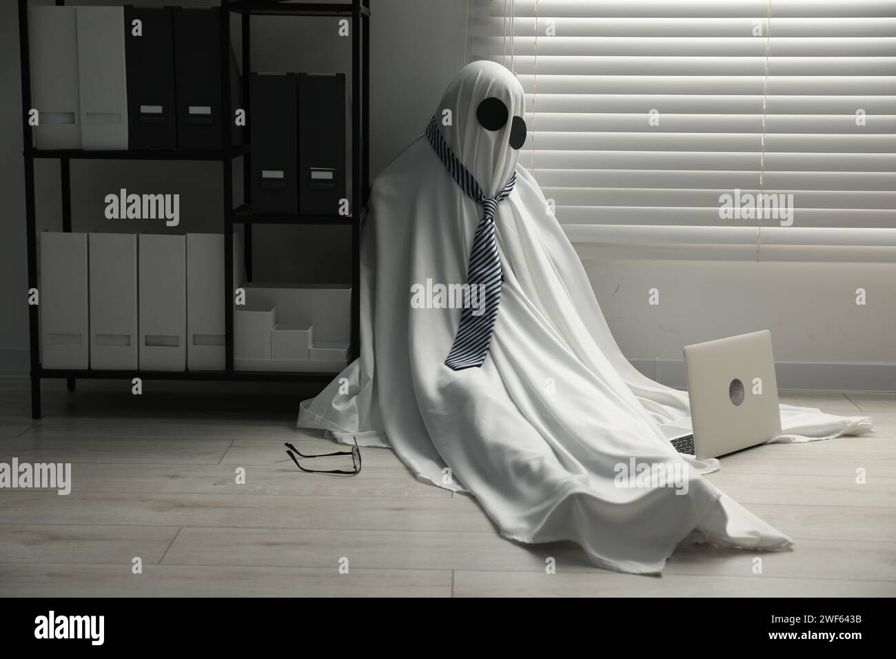 Overworked ghost. Man in white sheet with laptop on floor in office ...