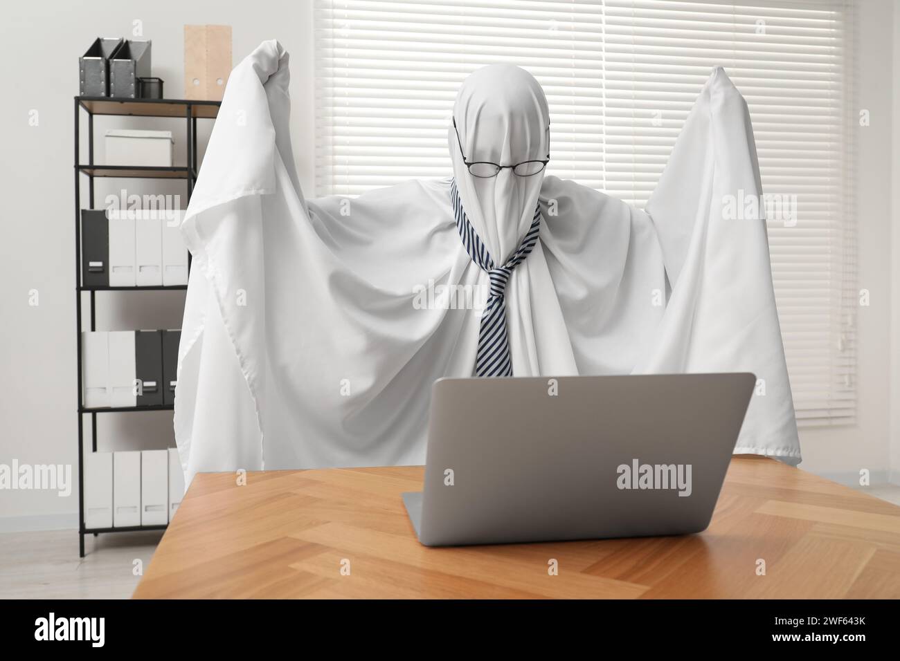 Overworked ghost. Man covered with white sheet using laptop at wooden ...