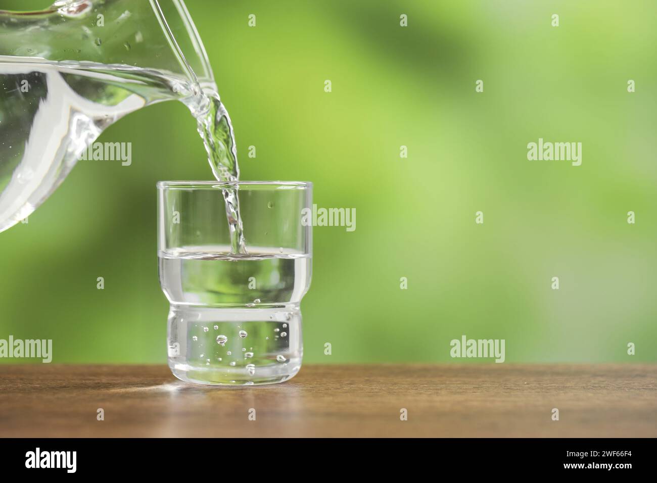 Pouring fresh water from jug into glass on wooden table against blurred ...