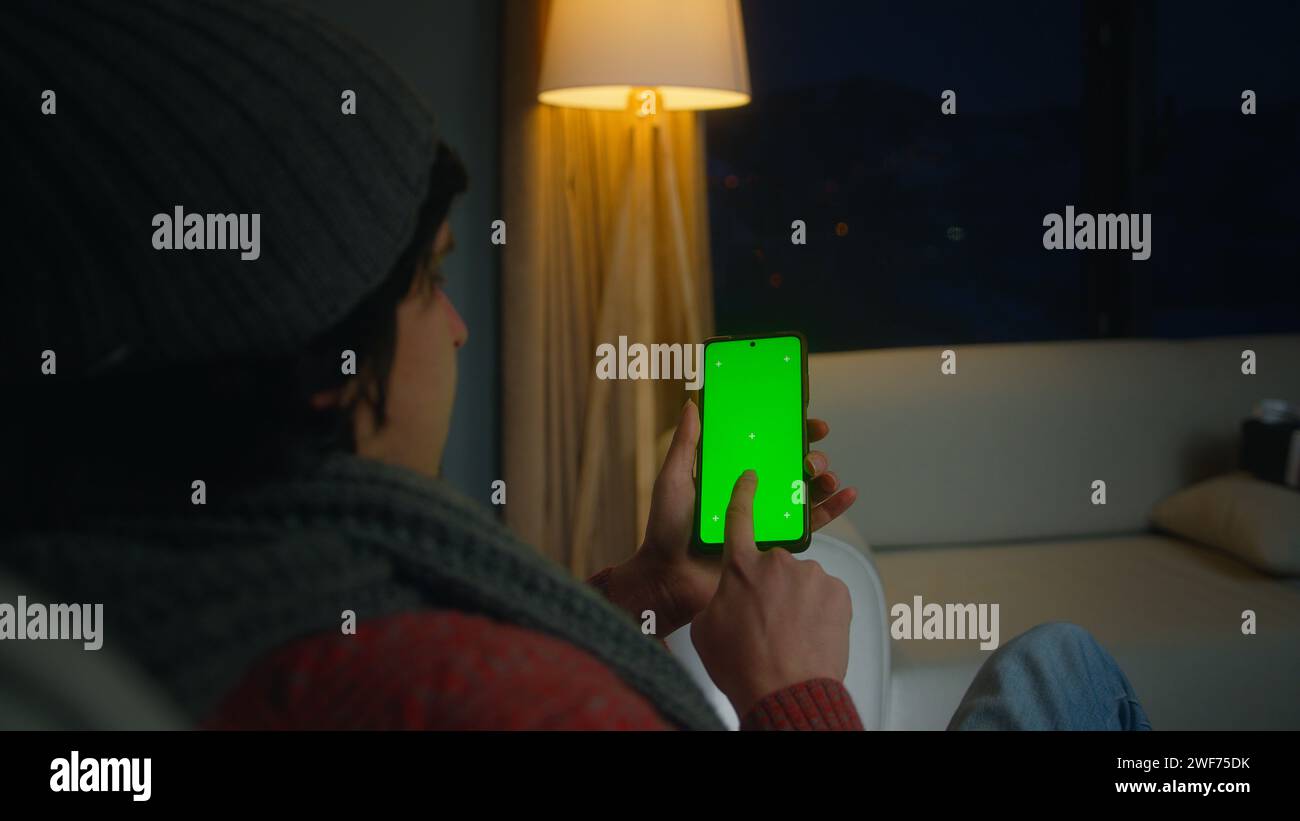 man using, tapping on screen smartphone with green mock-up screen. Male sitting on sofa ...