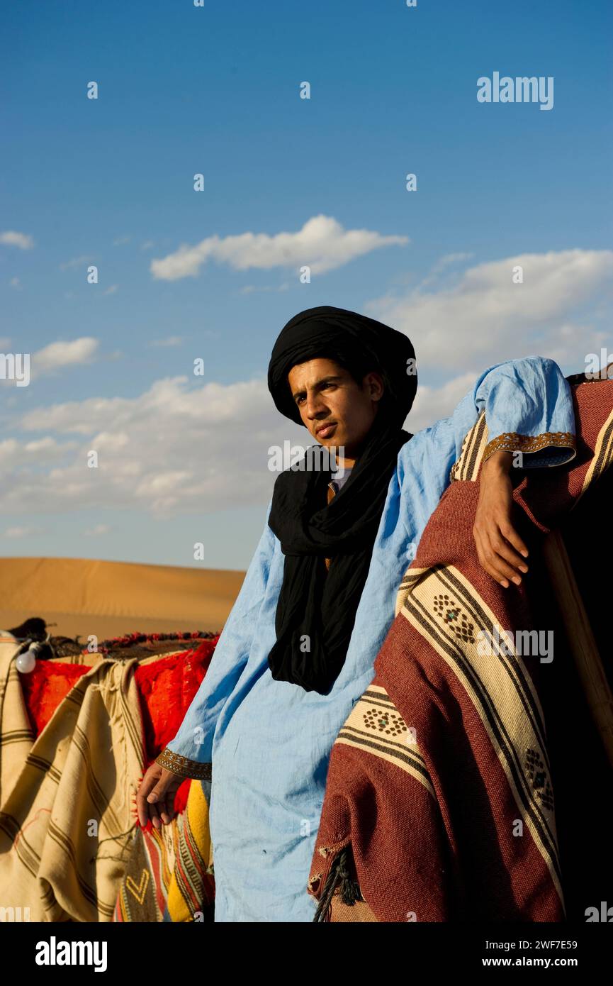 A desert guide in traditional dress stands in a tent camp with brightly ...