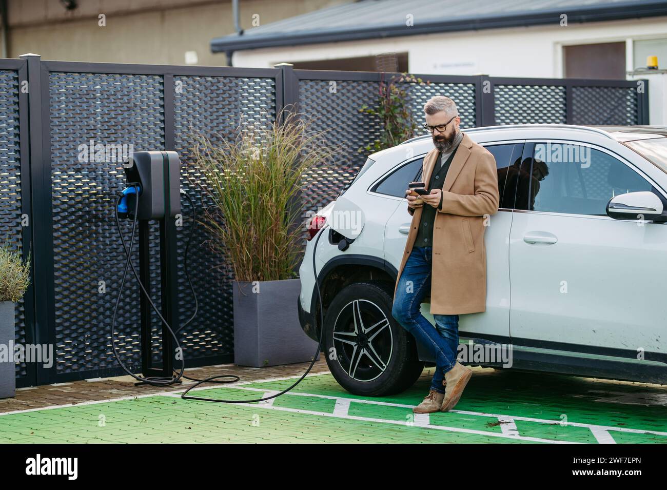 Portrait of businessman waiting while electric car is charging, leaning ...