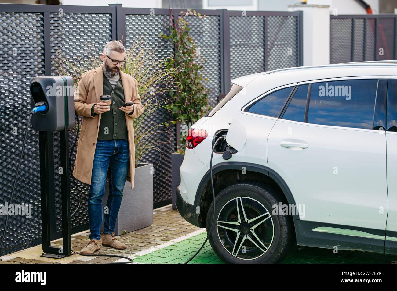 Portrait of businessman waiting while electric car is charging, leaning ...
